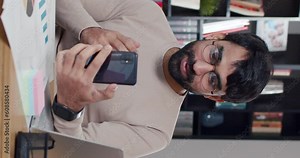 Vertical shoot. Adult man sitting at background of bookshelf and using phone for online mitting. Young buisinessman cooperate with colleaques via video call. Consept of online learning and work.
