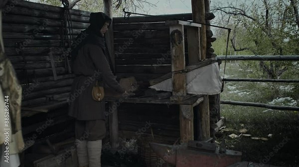 Full shot of young Caucasian male smith stirring coals in forge and fanning fire with bellows, while heating iron for beating with hammer on anvil, while at work in open-air wooden smithy