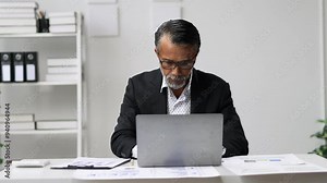 Focused businessman analyzing reports and working on a laptop in a modern office setting. Professional and productive, he is immersed in his work, managing documents and making notes.