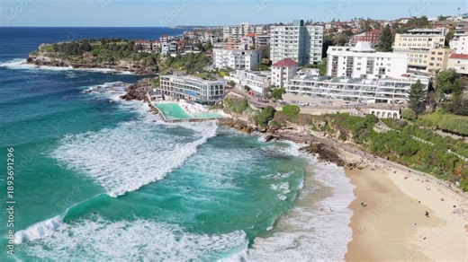 Epic aerial over waves crashing on the sand at Bondi Beach in Sydney, Australia, with scenic Pacific views of cliffs and hotels in the background as surfers, swimmers and tourists enjoy the beach.
