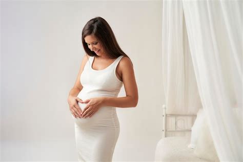Una mujer embarazada con un vestido blanco formando un corazón con las