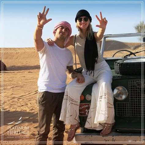 Two People Sitting On Top Of A Green Car In The Desert With Their Hands Up