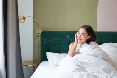 Beautiful Sleepy Asian Girl Wakes Up In Her Bed With Cozy White Blankets Looking Outside Window