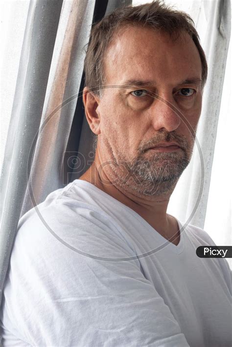 Image Of 50 55 Year Old Caucasian Man In A White T Shirt Standing By A Window With A Serious