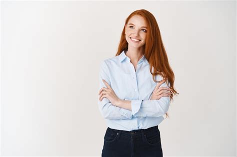 생강 머리와 파란 눈을 가진 웃는 여학생은 왼쪽 상단 모서리에서 희망적으로 웃고 가슴에 팔짱을 끼고 흰 벽에 파란색 칼라