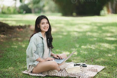 Asian Woman Sitting On Picnic Blanket And Lawn At Park Working On
