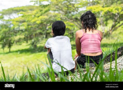Two Brunette Latina Girls Sitting Next To A Tree In A Tall Grass Field Admiring Nature Stock