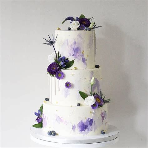 A Three Tiered White Cake With Purple Flowers On The Side And Green