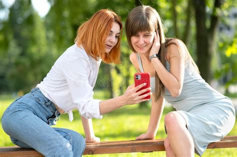 Two Young Girls Friends Sitting On A Bench In Summer Park Having Conversation On A Mobile Phone