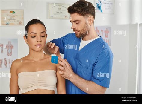 Handsome Bearded Doctor Putting Kinesiological Tapes On Shoulders Of His Young Female Patient