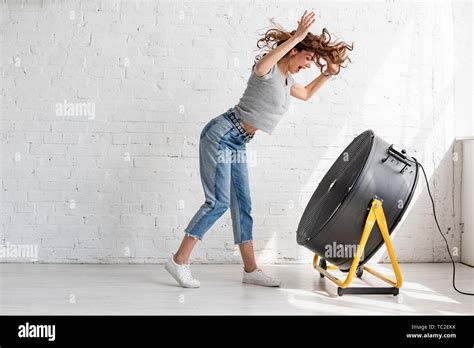 Excited Young Woman In Blue Jeans Standing With Raised Hands In Front Of Blowing Ventilator