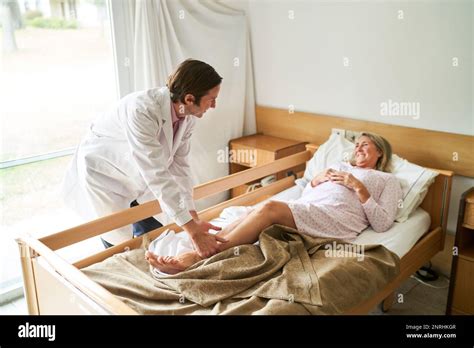 Smiling Male Doctor Lifting Legs Of Elderly Female Patient Lying In Bed