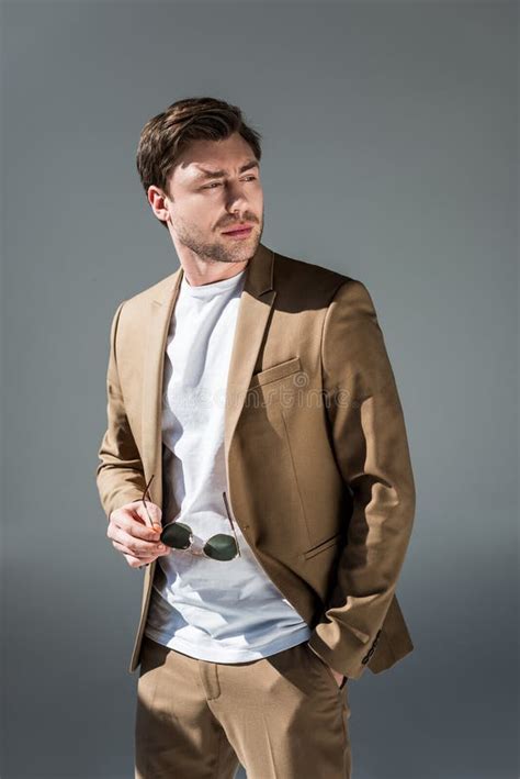 Handsome Man In Beige Suite Holding Sunglasses And Looking Away On Grey