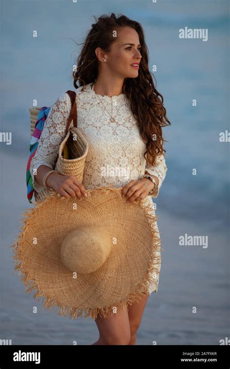 Modern Woman In White Dress And Straw Hat On The Seashore At Sunset