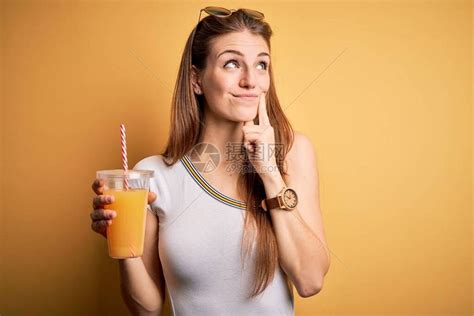 年轻美丽的红发美女在黄色背景下喝着健康橙汁面对严肃的面孔思考问题高清图片下载 正版图片504639662 摄图网