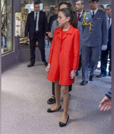 A Woman In An Orange Coat And Black Shoes Standing Next To A Man In A Suit