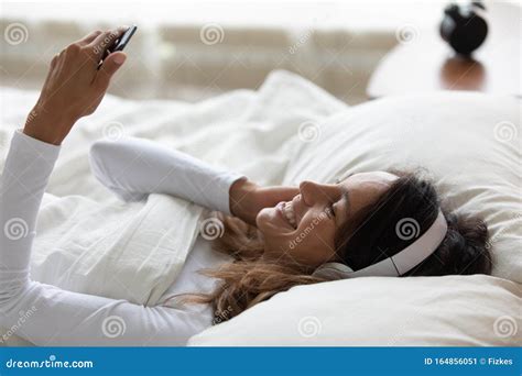 Woman Lying In Bed Listening Music Using Phone And Headphones Stock