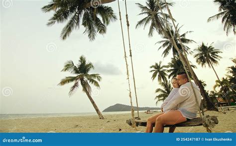 Loving Couple On The Swing On Tropical Beach Woman And Man In White Shirt Enjoy Gold Sunset And