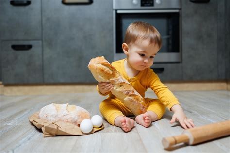 Un lindo niño de un año está sentado en la cocina y comiendo un pan