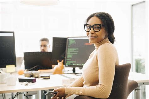 Portrait Of Female Programmer Sitting By Desk With Colleague In