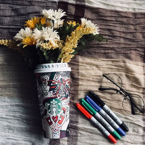 Flowers In A Starbucks Cup With Markers And Eyeglasses Next To It On A Bed