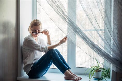 Barefoot Woman In Glasses And White Shirt Sitting On The Windowsill