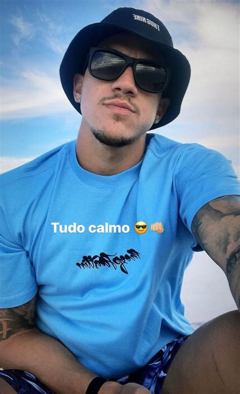 A Man Wearing Sunglasses And A Hat Sitting On The Beach With His Hands