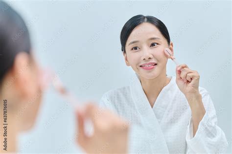 아시아 한국의 검은머리의 젊은 여자 모델이 밝은 표정으로 거울잎에 앉아 샤워가운을 입고 거울을 보며 핑크색 마사지 롤러로 얼굴