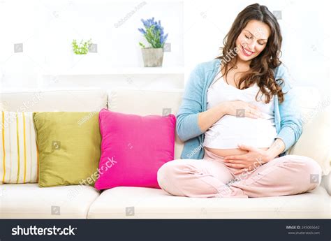 Pregnant Happy Smiling Woman Sitting On A Sofa And Caressing Her Belly
