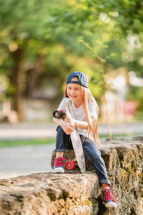 Girl In Cap Sits Backwards On Stone Holds Skateboard With Her Hands On Her Knee Stock Image