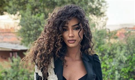 A Close Up Of A Person Wearing A Black Shirt And Curly Hair With Trees