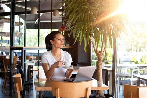 Young Beautiful Girl Sitting At A Table In A Cafe By The Window Drinking Cappuccino Coffee