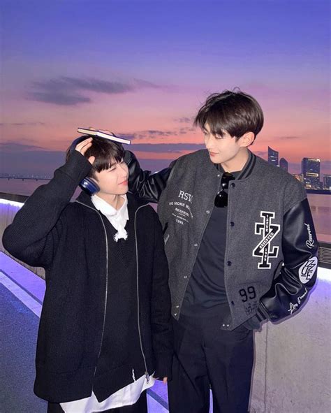 Two Young Men Standing Next To Each Other Near The Ocean At Sunset With City Lights In The