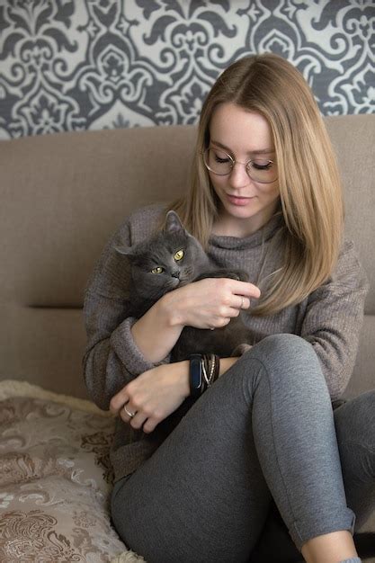 Premium Photo Beautiful Girl Sits At Home On The Sofa And Hugs Her Gray Cat With Bright Yellow