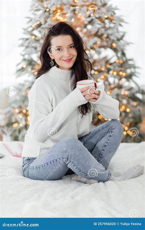 A Beautiful Brunette Girl In A White Sweater And Blue Jeans Sits On A Bed With A Cup Of Hot