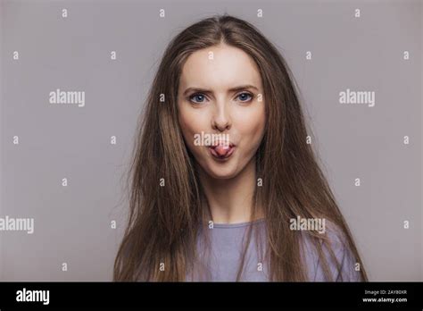Funny Brunette Female With Atractive Look Shows Tongue Makes Grimace And Has Fun Indoor Looks